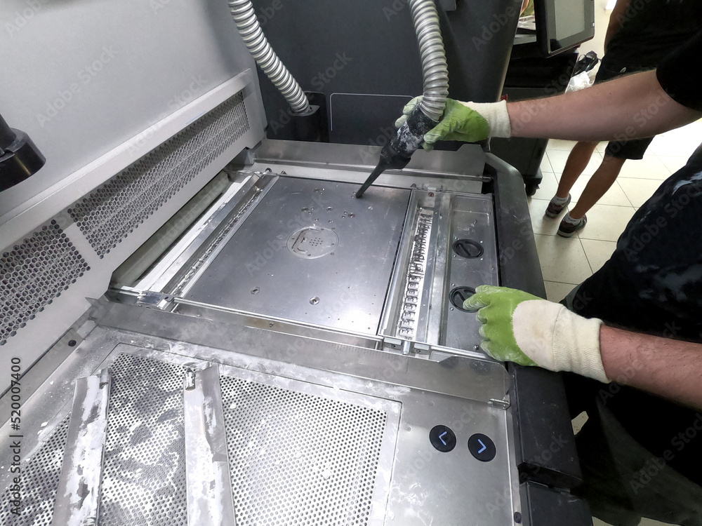 A male worker cleans the surface of an industrial 3D printer from white ...