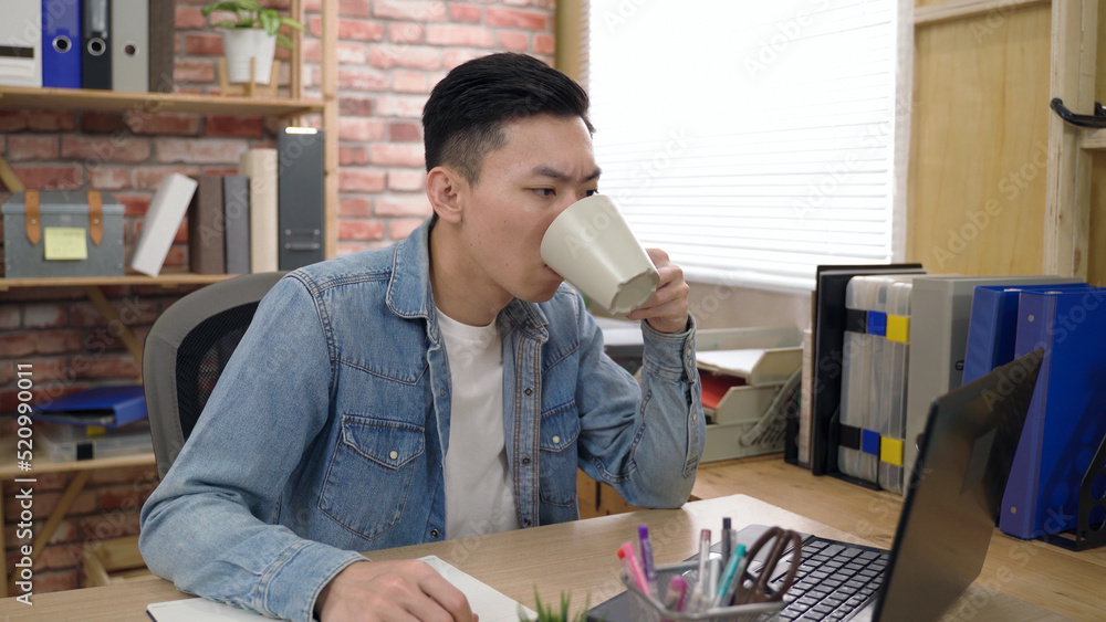 asian creative worker pausing to sip tea and fold his arms at desk is ...