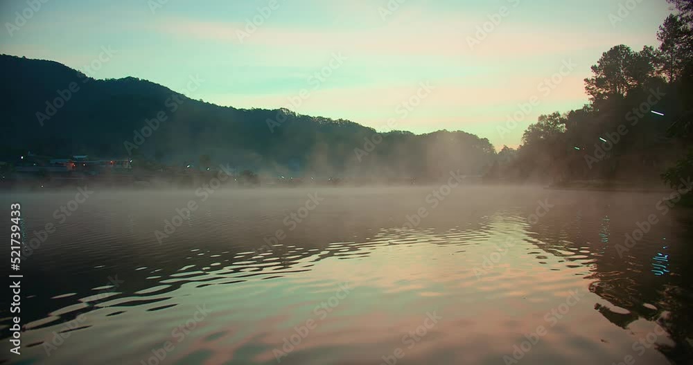 Dawn breaks over a tranquil lake shrouded in mist, with a lush mountain ...
