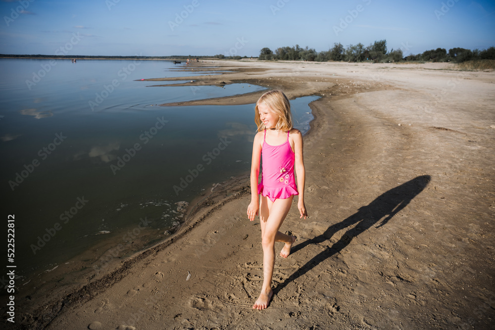 A little seven-year-old girl in a pink swimsuit runs along the lake in ...