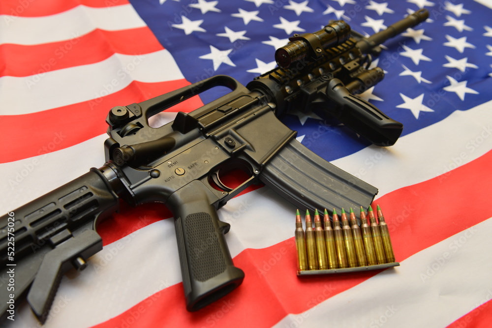 A closeup of an automatic carbine rifle over the US flag with stacks of ...