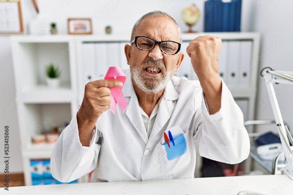 Mature doctor man holding pink cancer ribbon at the clinic annoyed and ...