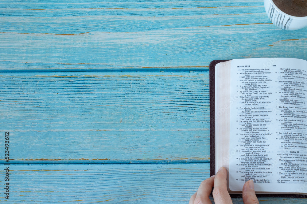 Human hand holding open Holy Bible Book with a cup of coffee on a blue wooden background with ...