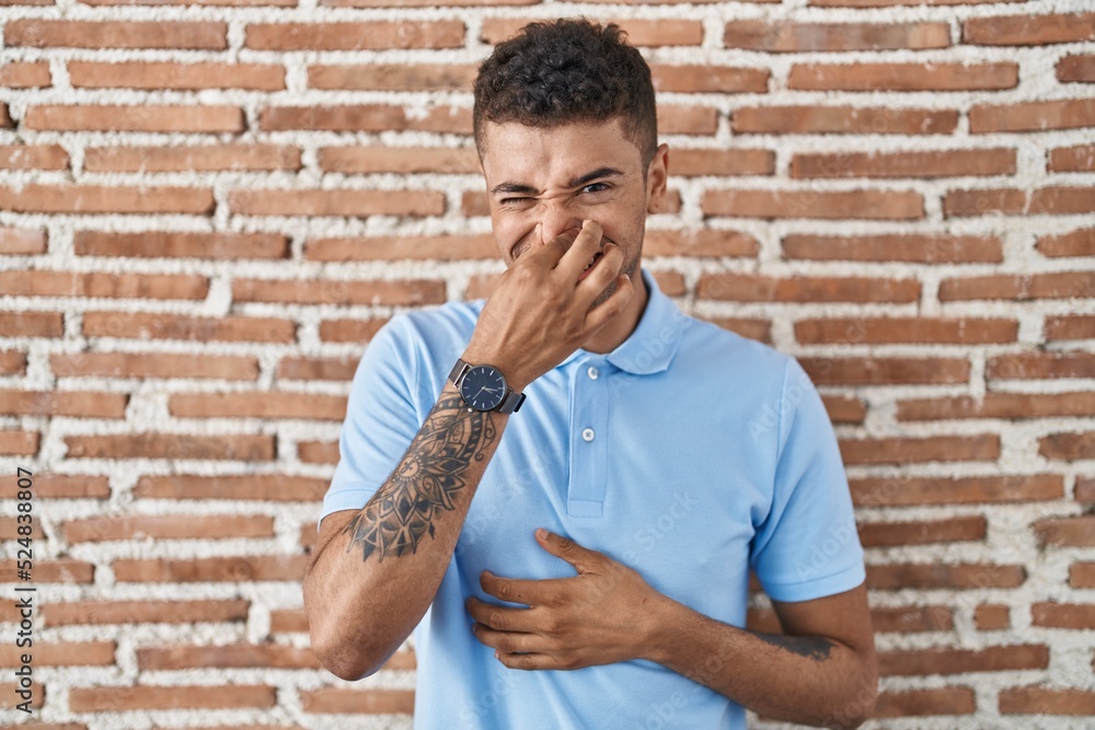 Brazilian young man standing over brick wall smelling something stinky ...