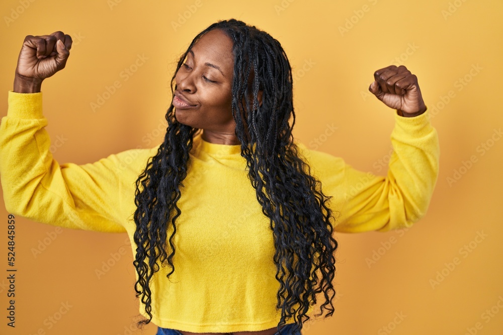 African woman standing over yellow background showing arms muscles ...