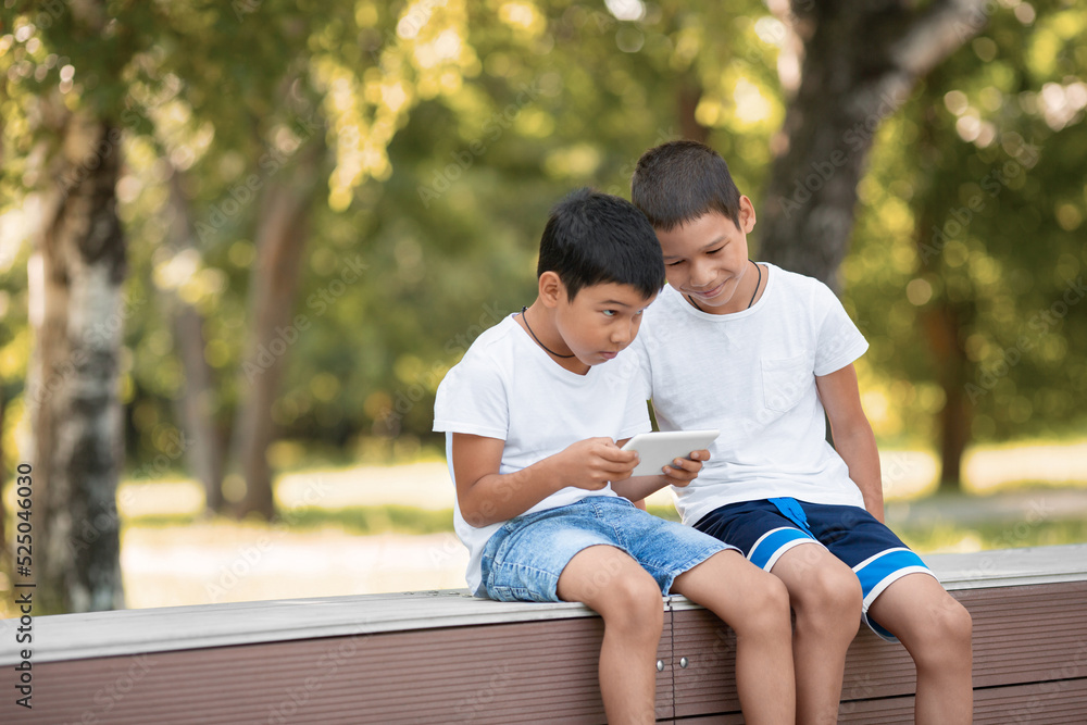 Two boys using together a digital tablet sitting outdoor in park in ...