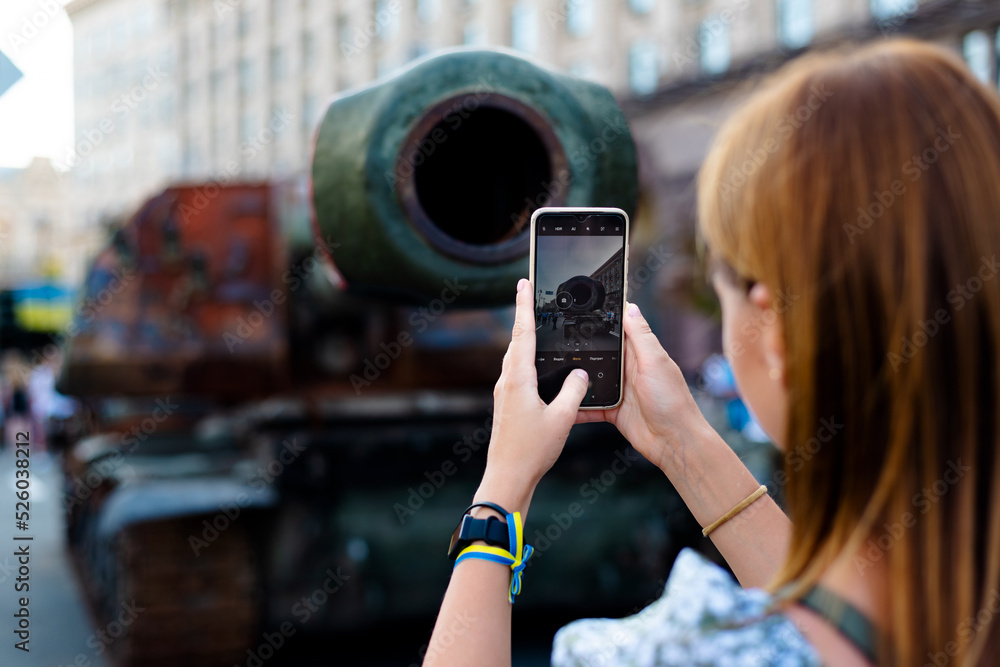 girl takes a photo on the phone of a burnt Russian tank on the street ...