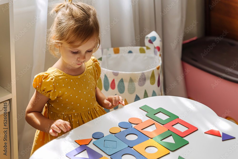 A little girl playing with wooden shape sorter toy on the table in playroom. Educational boards ...