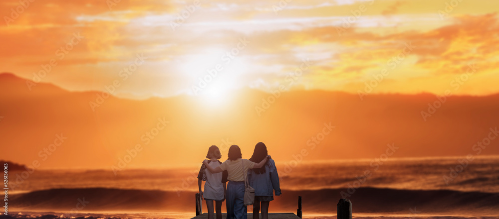 three women stand on the beach pier hugging back view sunset sky ...