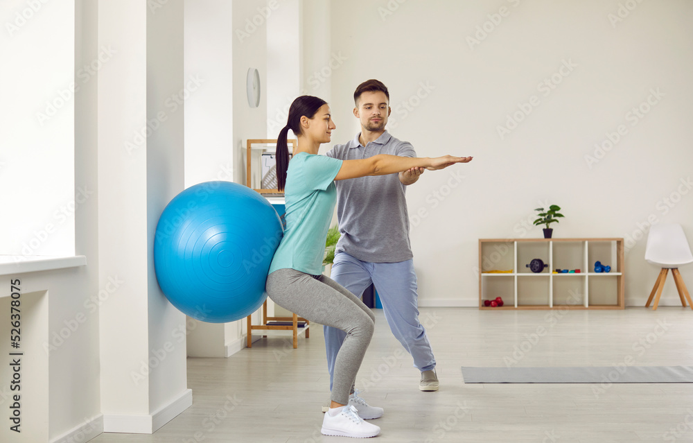 Fitness trainer helping young female patient do wall squats with blue ...