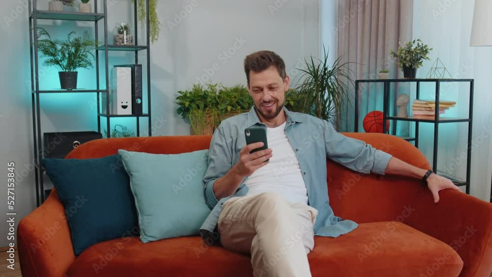 Portrait of adult man sitting on sofa uses mobile phone smiles at modern home apartment. Young ...