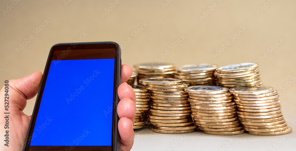 Cell phone with blue screen in hand and background with stacks of coins ...