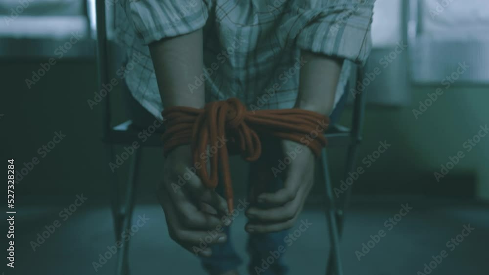 A kidnapped young woman tied is sitting on chair. Hands tied with a rope. She's trying to free ...