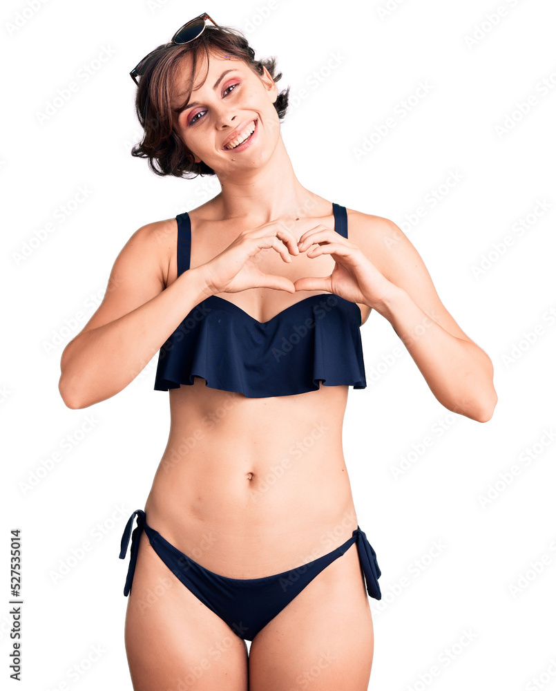Beautiful young woman with short hair wearing bikini smiling in love showing heart symbol and ...