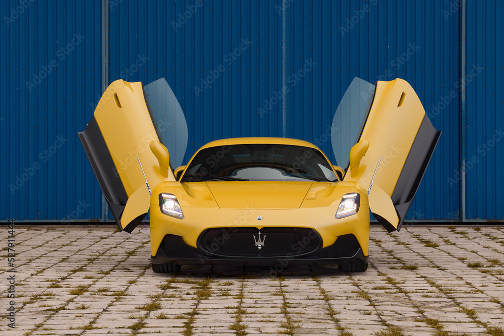 A yellow Maserati MC20 with an open door against a blue tin wall. Front ...