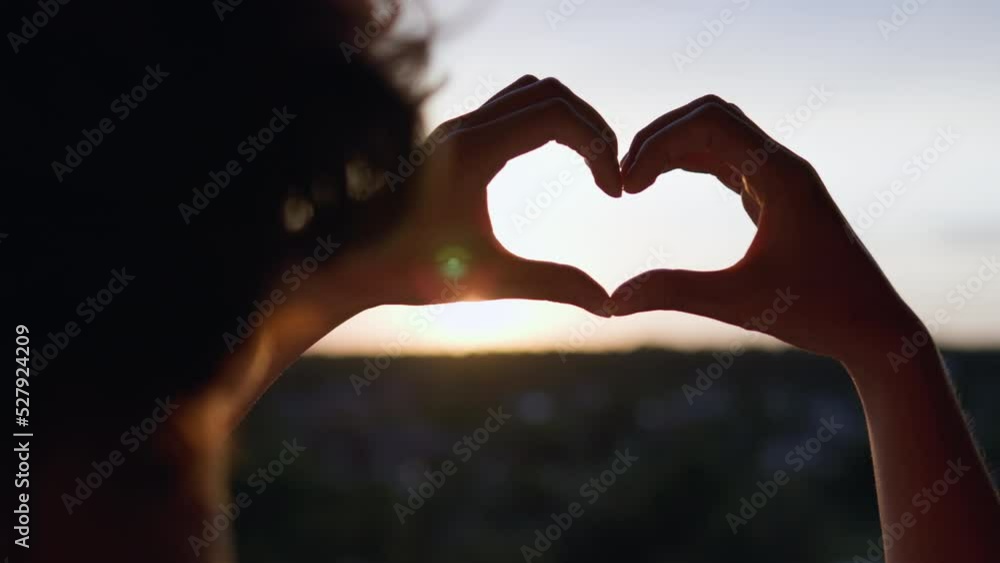The girl stands in front of the sun and shows hearts with her hands ...