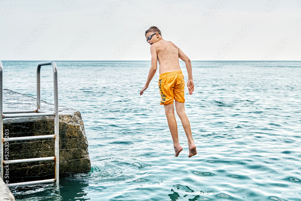 Moments of schoolboy jumping from stone pier with ladder into sea doing ...