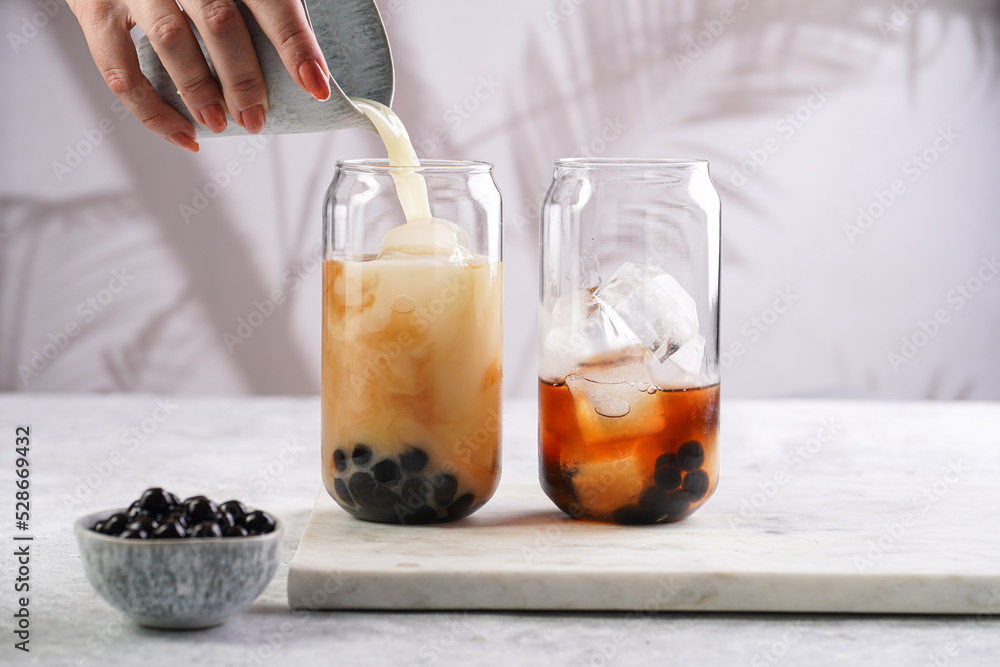 Female hand pouring milk into glass with black tea, ice cubes and ...