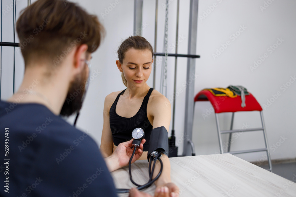 Male doctor measures the pressure of a young sports woman in the gym ...