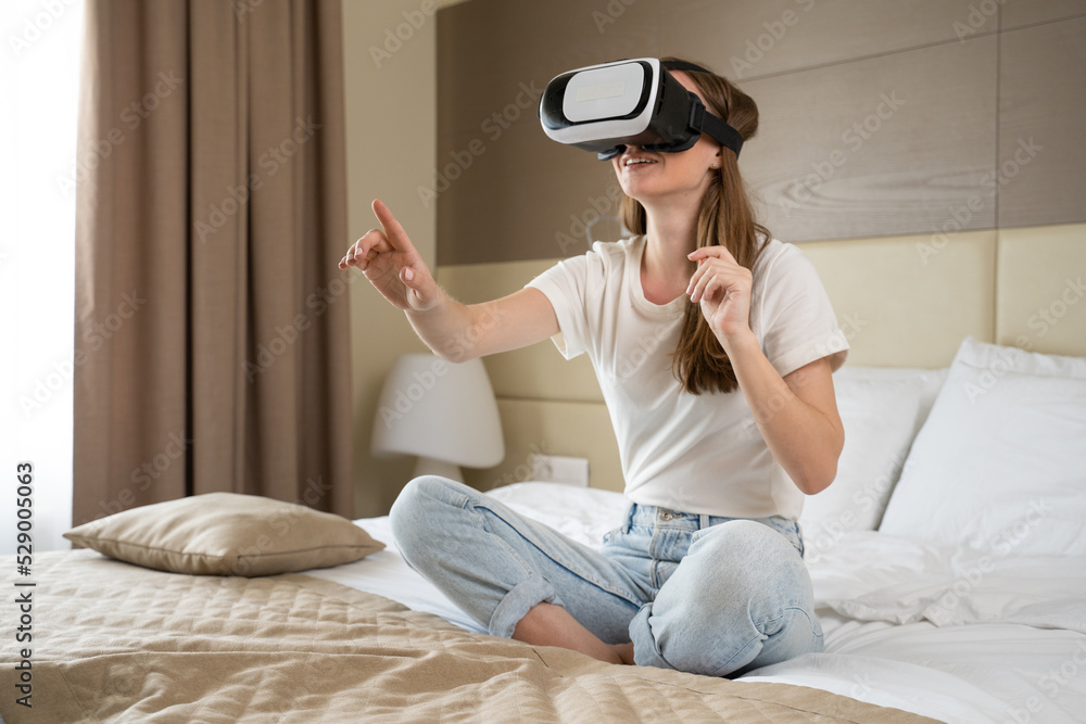 Excited woman sitting on king-size bed plays VR games in hotel room ...