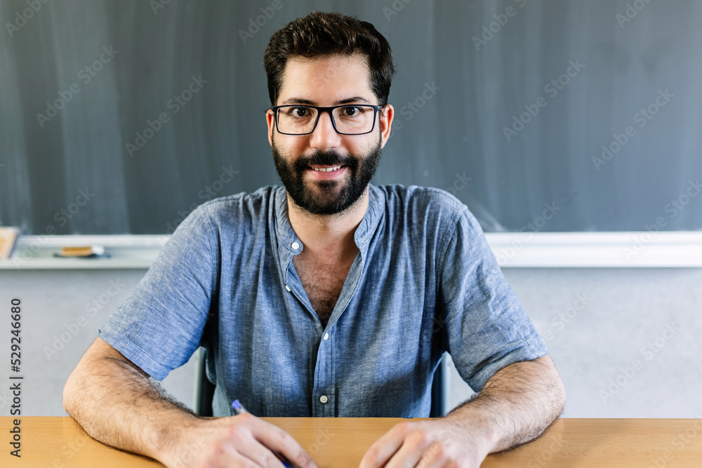Cheerful male teacher sitting at desk in classroom - Screen view face ...