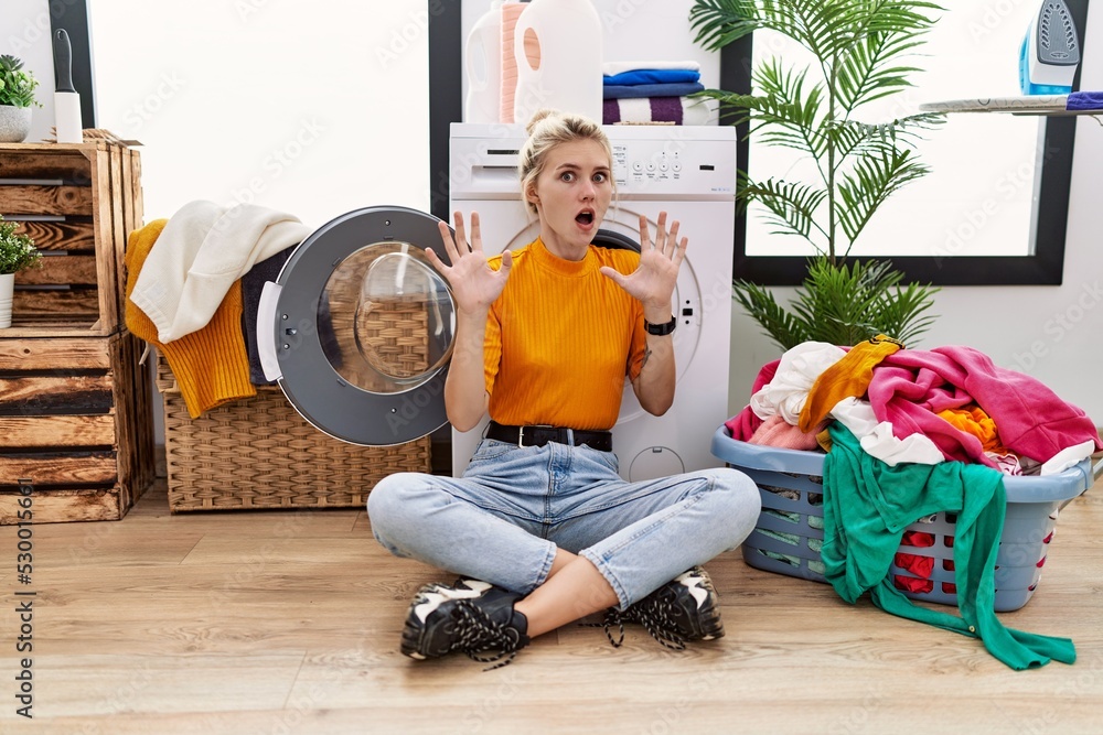 Young blonde woman doing laundry sitting by washing machine afraid and ...