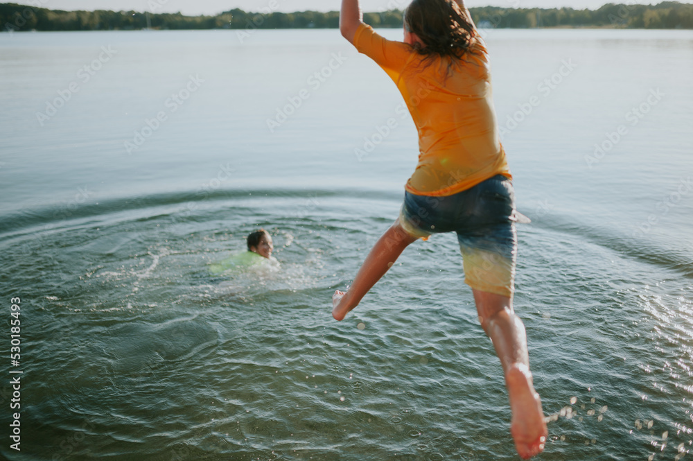Boy jumping off dock as another boy smiles swimming in the water Stock ...