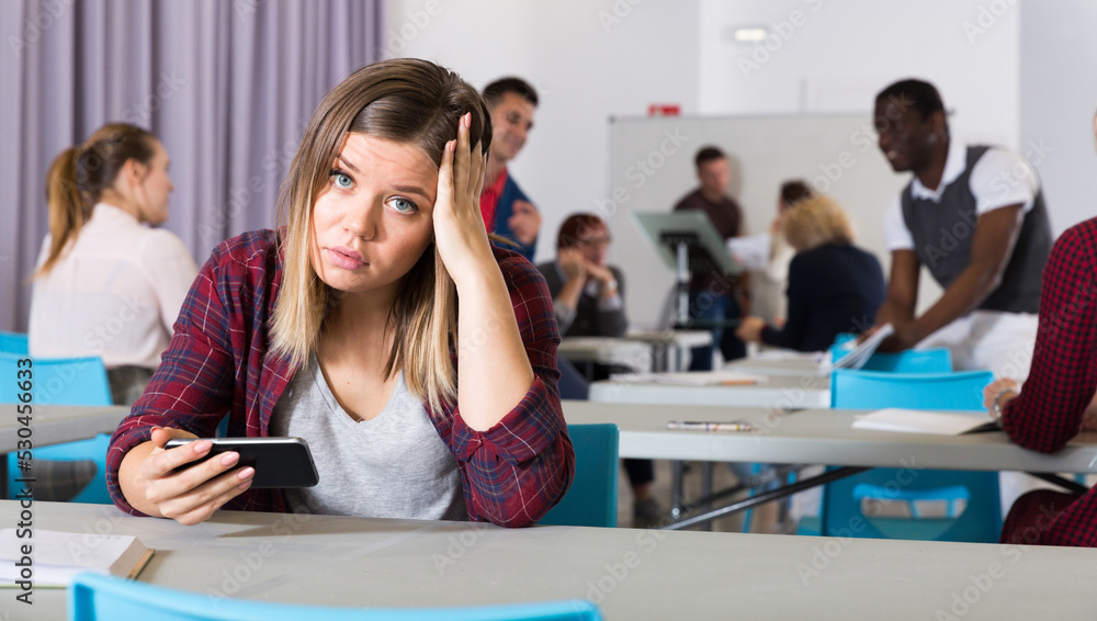 Frustrated young woman student sitting with phone in classroom in break ...
