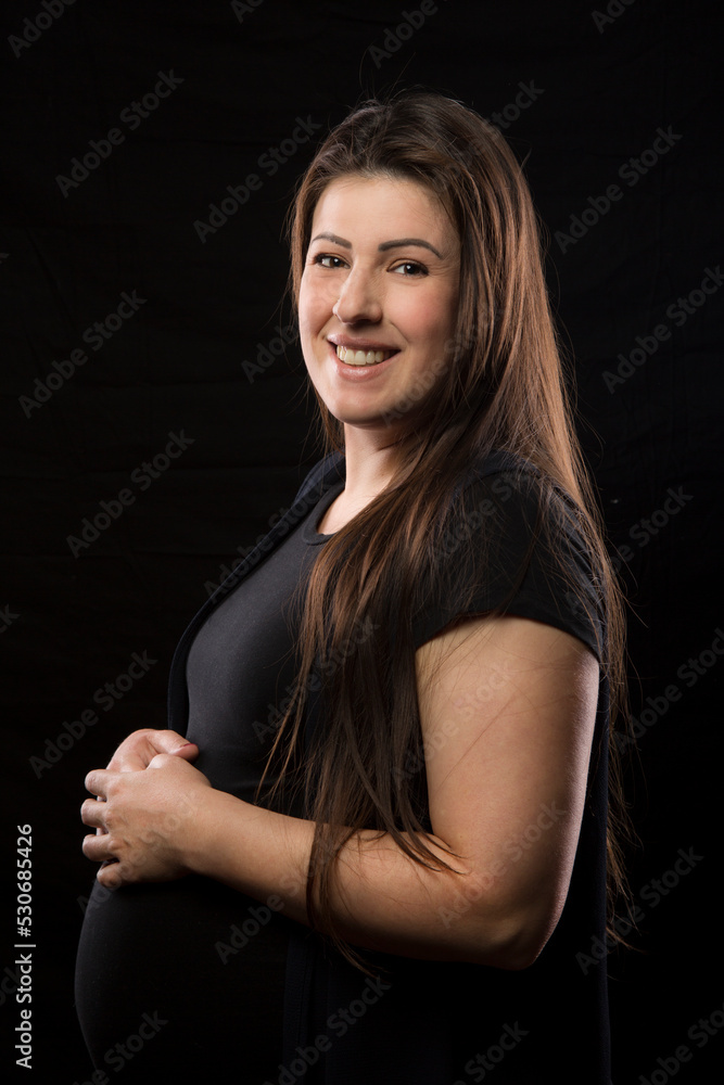 Young brunette woman at late stages of pregnancy smiles and poses with her baby bump, studio ...