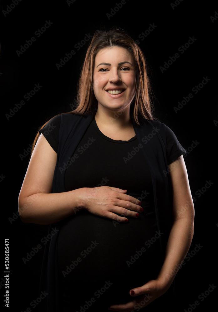 Young brunette woman at late stages of pregnancy smiles and poses with her baby bump, studio ...