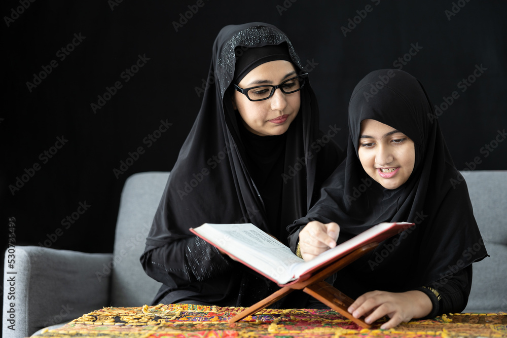 muslim mother with her daughter reading a holy book or Quran on sofa ...