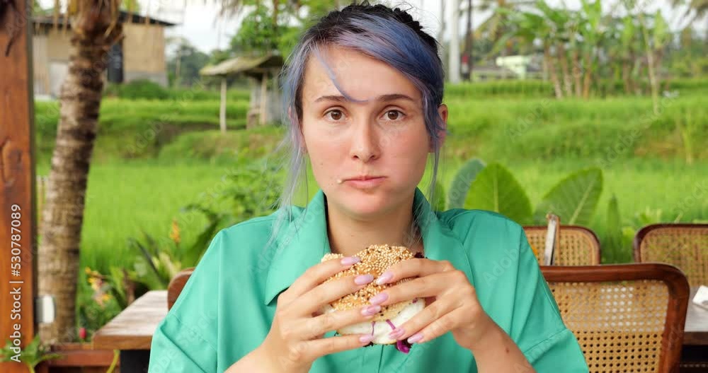 Pretty cheeky young woman with blue hair eating a big beef burger ...