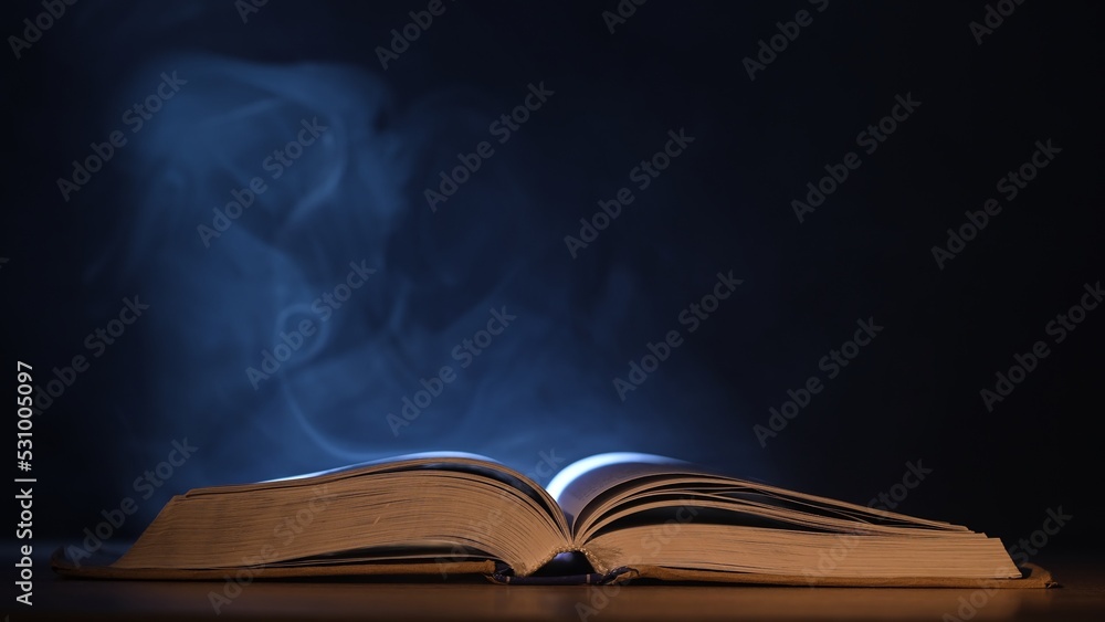 An open book illuminated by beam of blue light with swirling smoke on ...