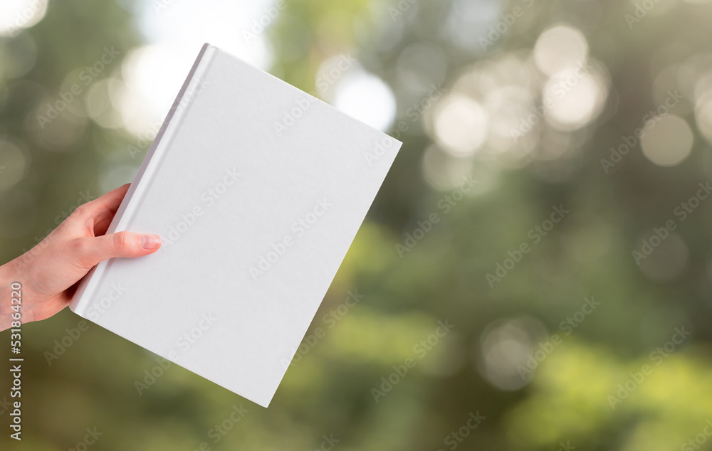 Hand holding book mockup on blurred background with green trees, grass ...