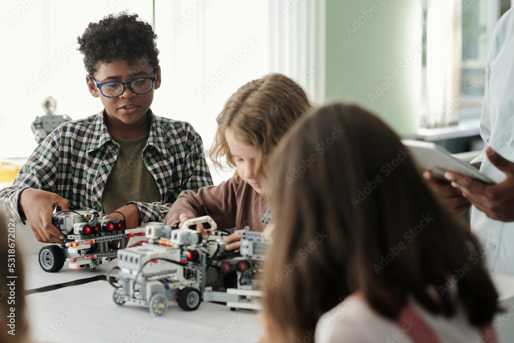 Cute intercultural schoolkids playing with toy robots which they ...