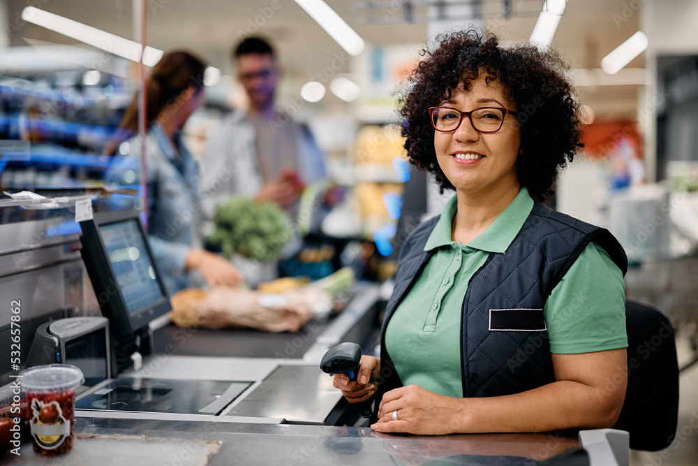 Happy woman working as cashier at supermarket checkout and looking at ...