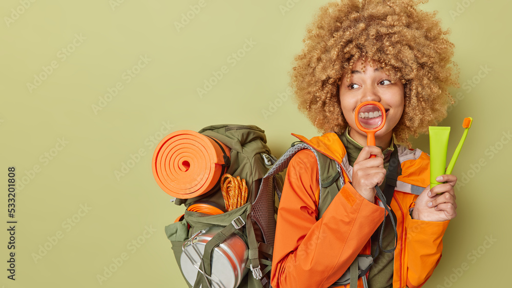 Positive curly female hiker backpacker holds magnifying glass over ...