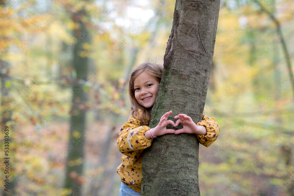 Smiling cute girl hugging tree and gesturing heart shape in forest ...