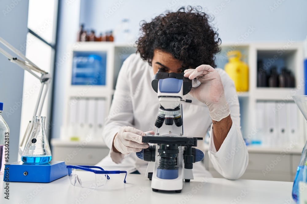 Young hispanic man wearing scientist uniform using microscope at ...