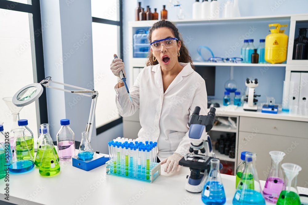 Young hispanic woman working at scientist laboratory scared and amazed ...