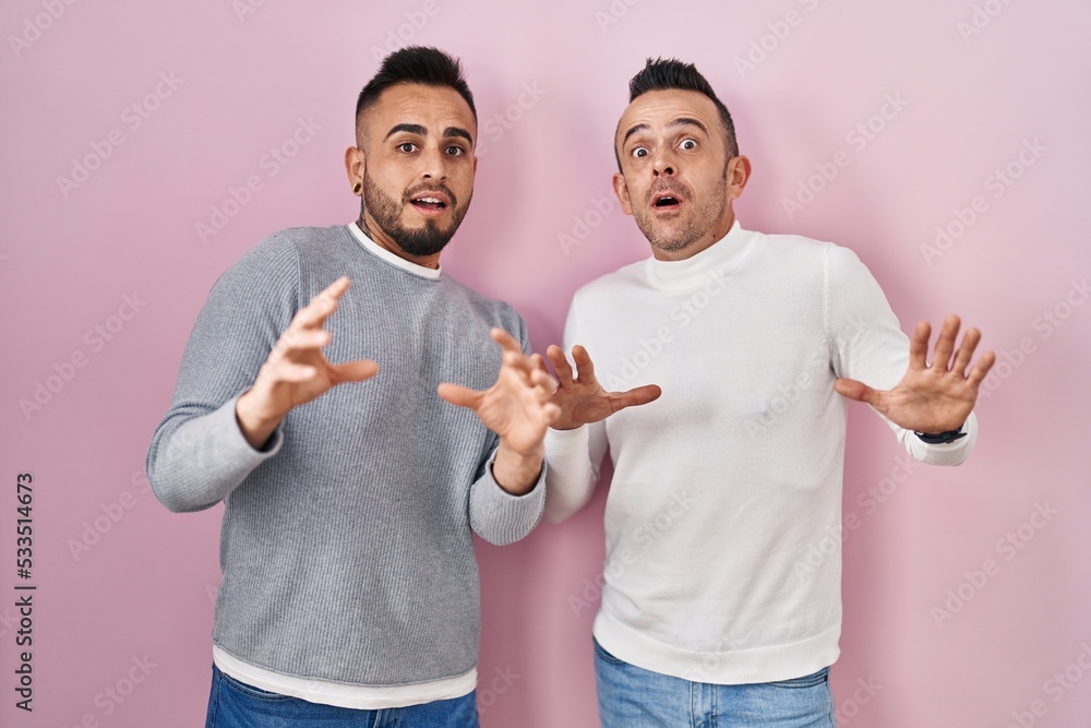 Homosexual couple standing over pink background afraid and terrified with fear expression stop ...