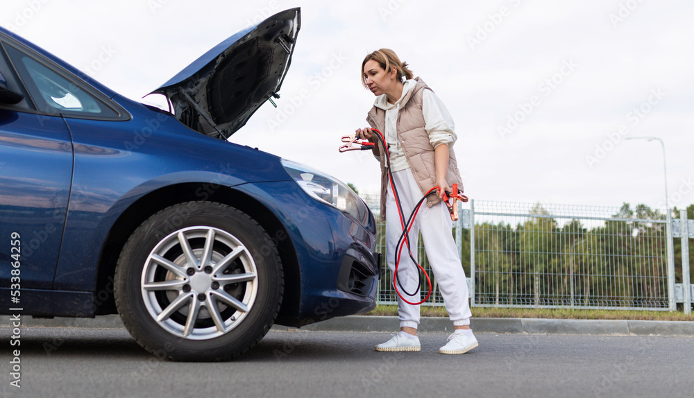 female driver at the open hood of a car with wires for charging the ...