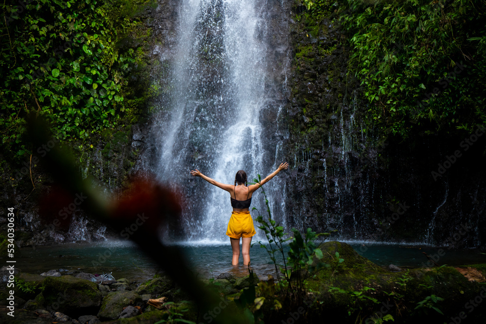 brave girl stands in front of a mighty waterfall with her hands raised ...