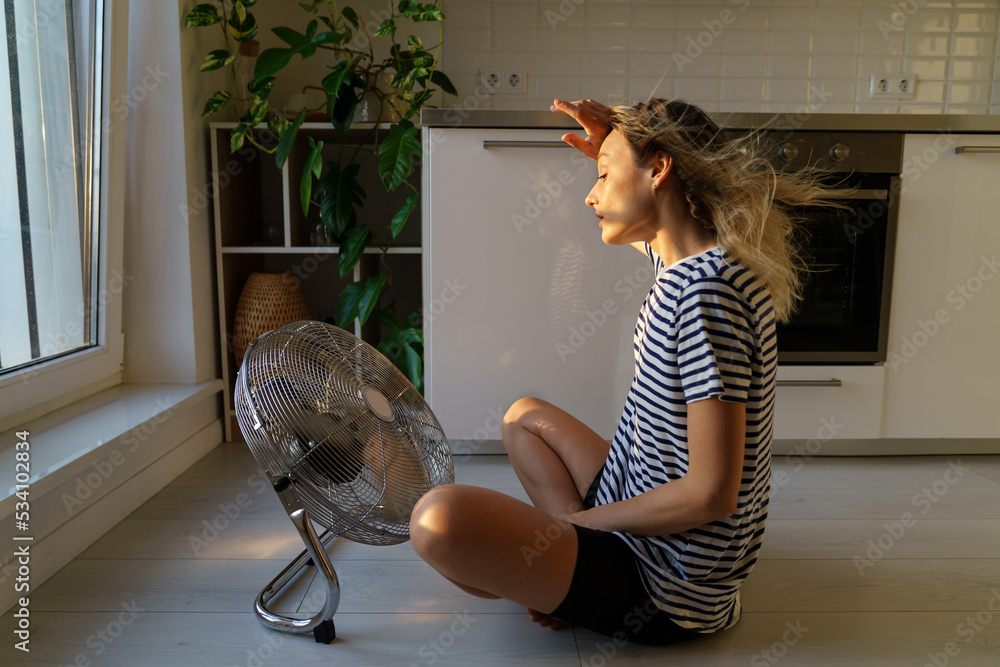 Exhausted young woman refreshing sit near big indoor ventilator blowing ...