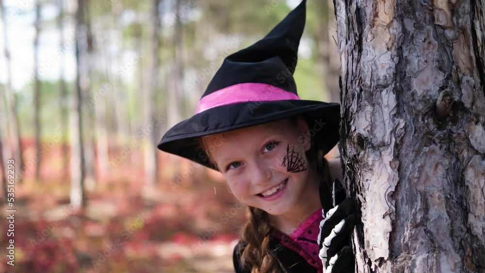 A girl dressed as a witch hides behind a tree in the forest. Halloween ...