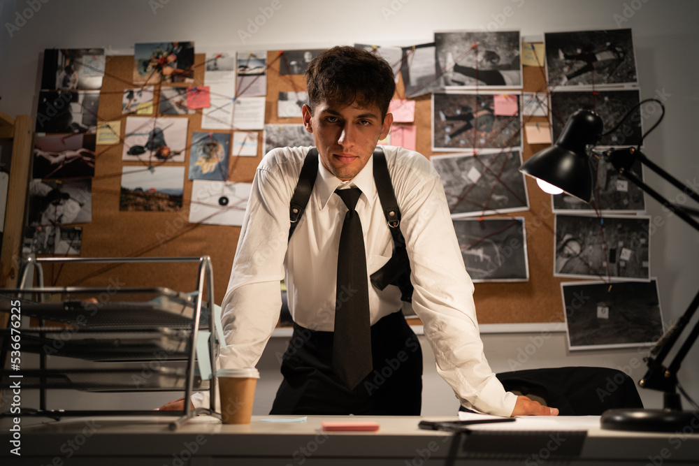 Young serious male detective in uniform bending over workplace with ...
