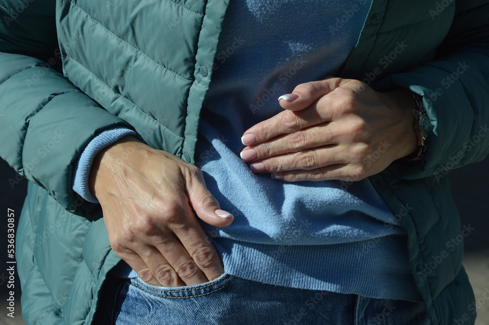 A woman in a casual sweater and jacket holds one hand on her stomach ...