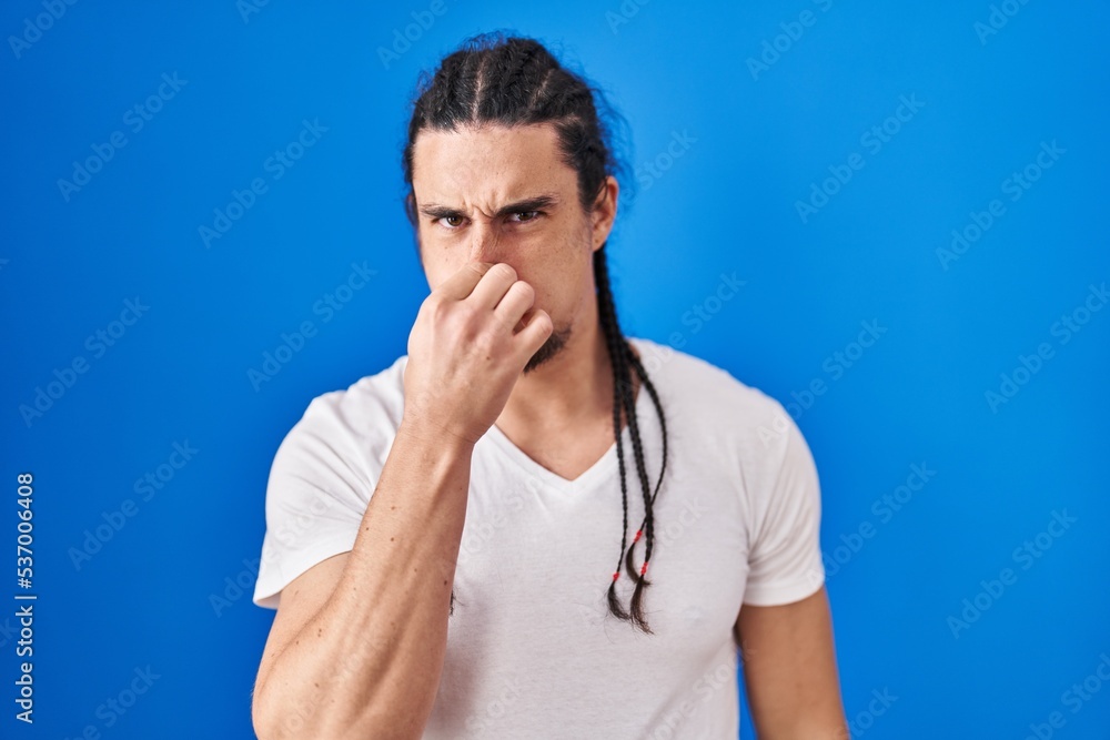 Hispanic man with long hair standing over blue background smelling ...