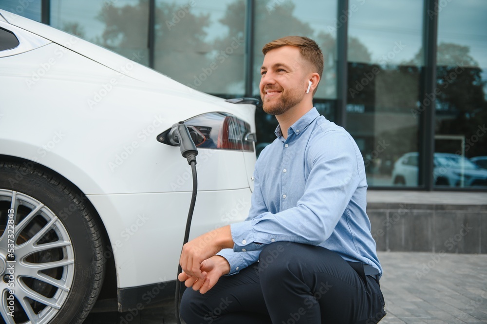 Hansome bearded guy sitting near his new modern electric car and ...