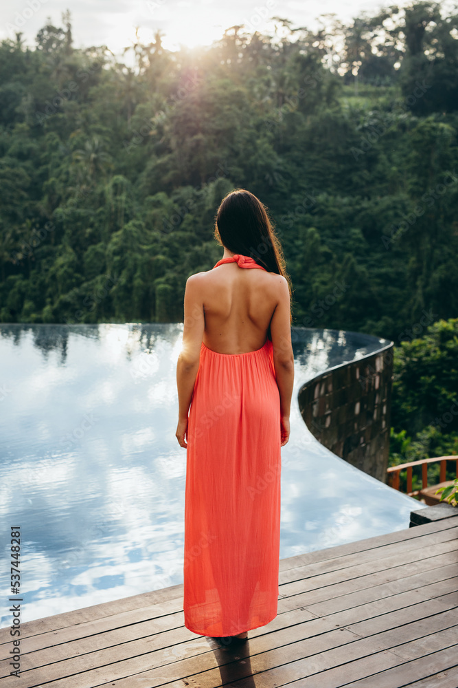 Rearview of a woman standing in front of an infinity pool in a sundress ...